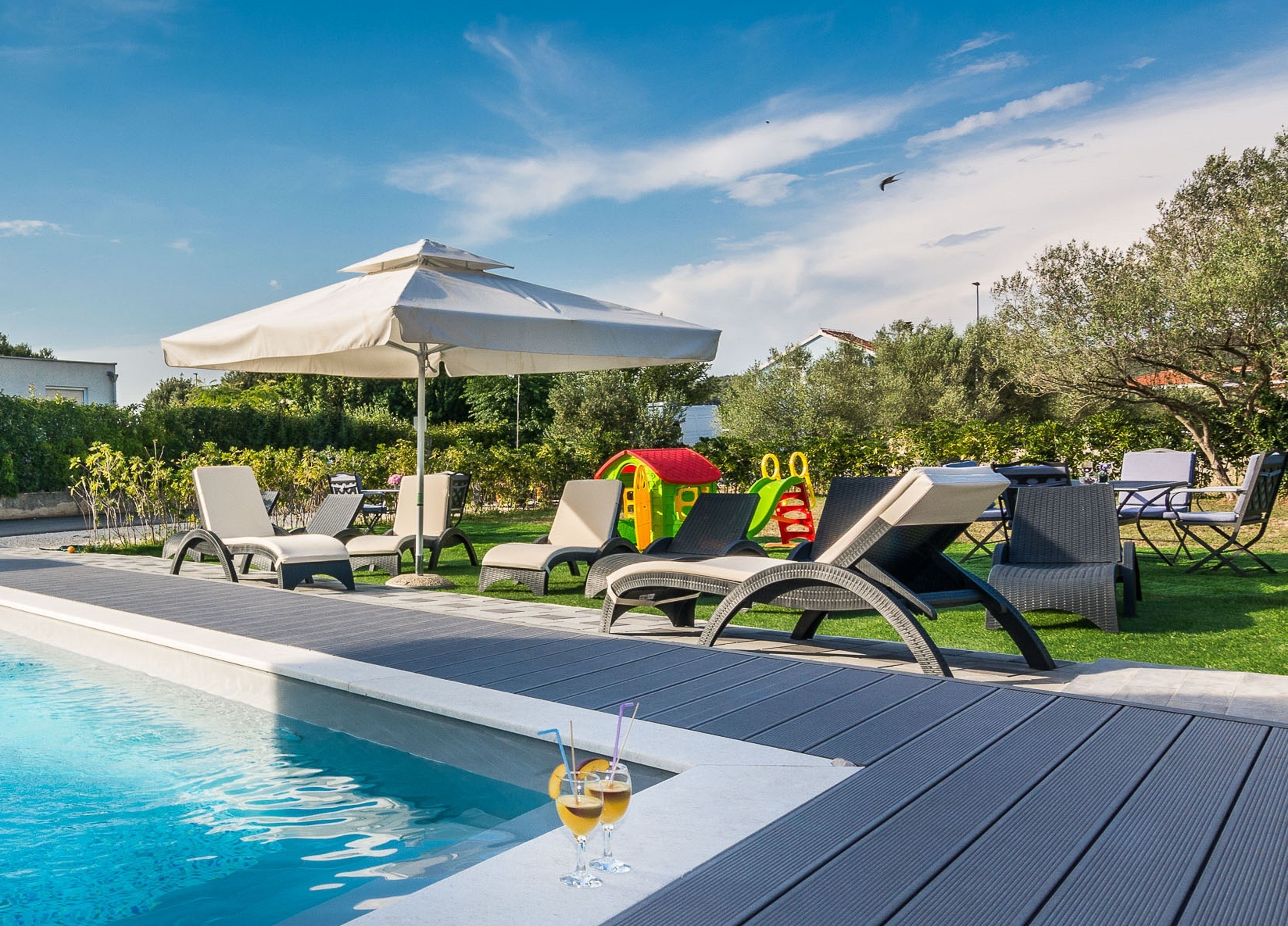 Uma área à beira da piscina com espreguiçadeiras, um guarda-sol e um parquinho infantil.