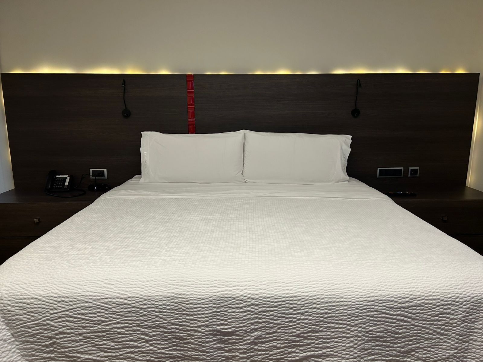 Uma cama bem arrumada com colcha branca, cabeceira escura com iluminação embutida e mesas de cabeceira com um telefone e um pequeno dispositivo eletrônico.