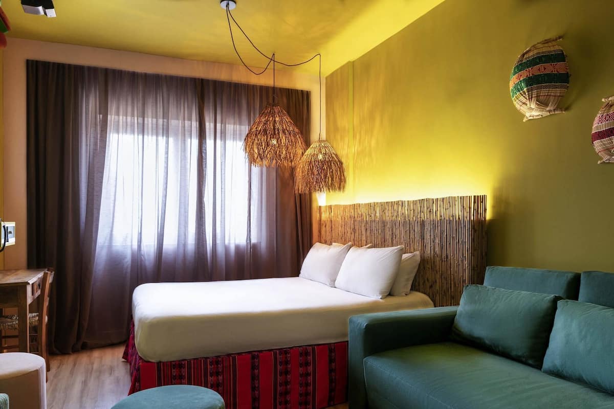 Quarto de hotel com cama, sofá verde, escrivaninha de madeira e uma grande janela com cortinas.