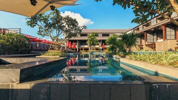 Piscina com reflexo límpido, rodeada por espreguiçadeiras e guarda-sóis, com um edifício ao fundo.