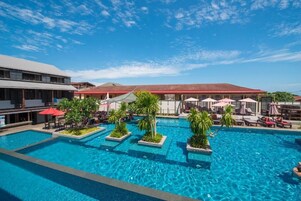 Piscina com várias pistas de natação, rodeada por espreguiçadeiras e guarda-sóis, adjacente a um edifício com telhado vermelho.