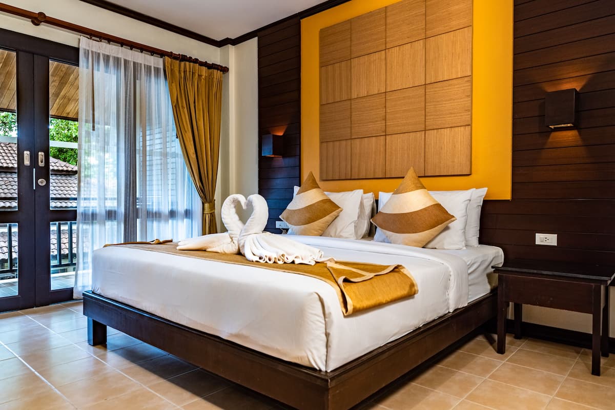 Quarto de hotel com uma cama grande, cabeceira de madeira, mesa de cabeceira pequena e vista para uma varanda.