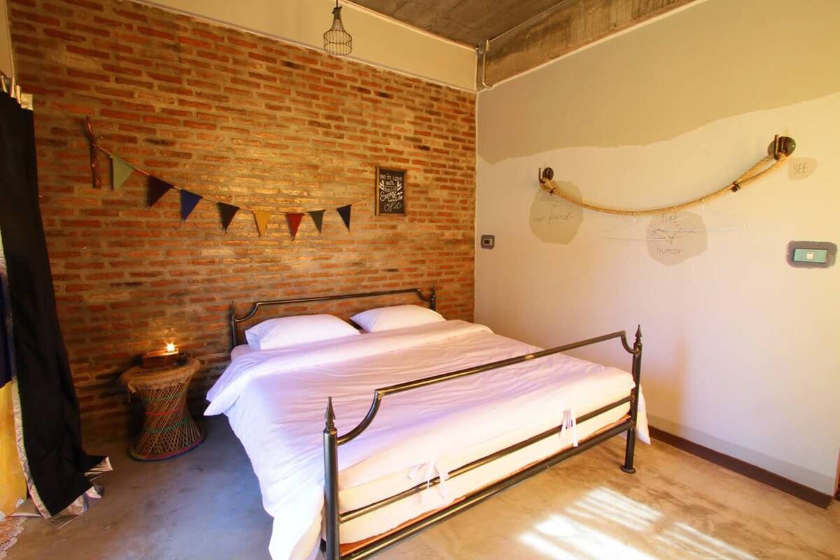 Quarto com estrutura de cama em metal, roupa de cama branca, parede de tijolos e balanço de corda fixado na parede.