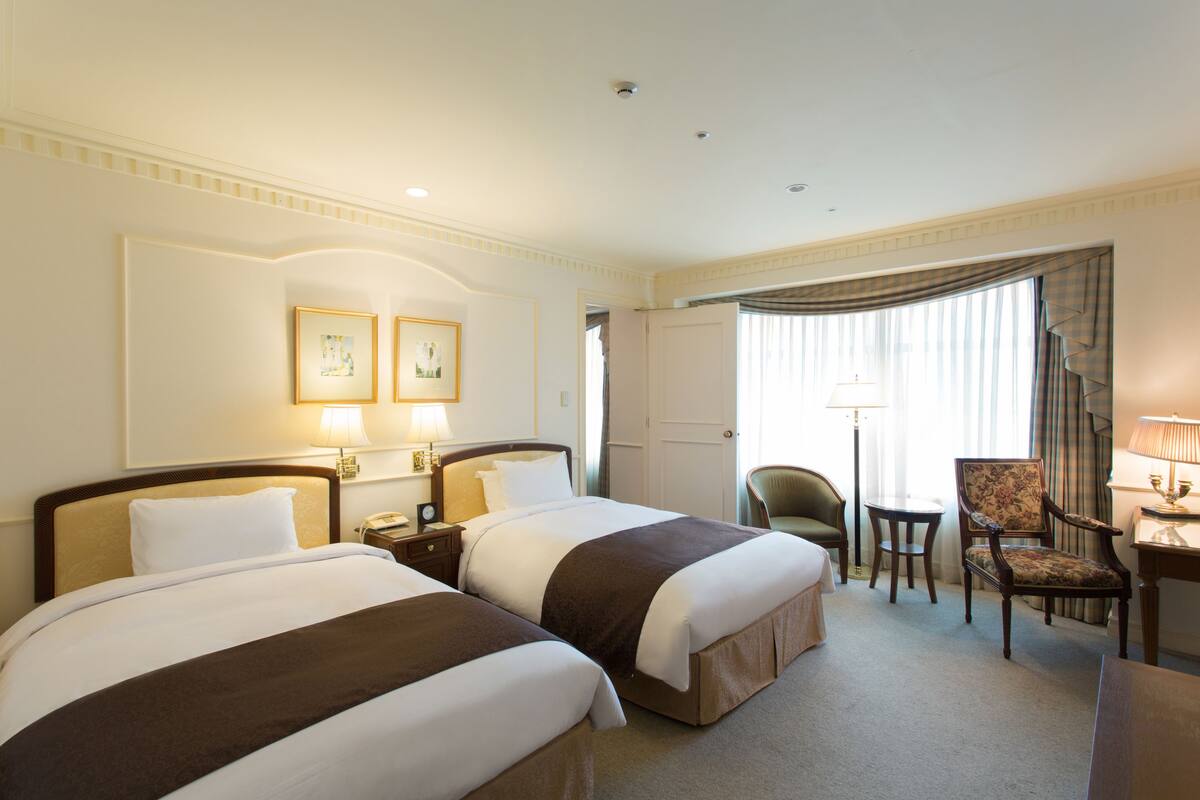 Quarto de hotel com duas camas, uma escrivaninha, uma cadeira e porta que dá para uma varanda.
