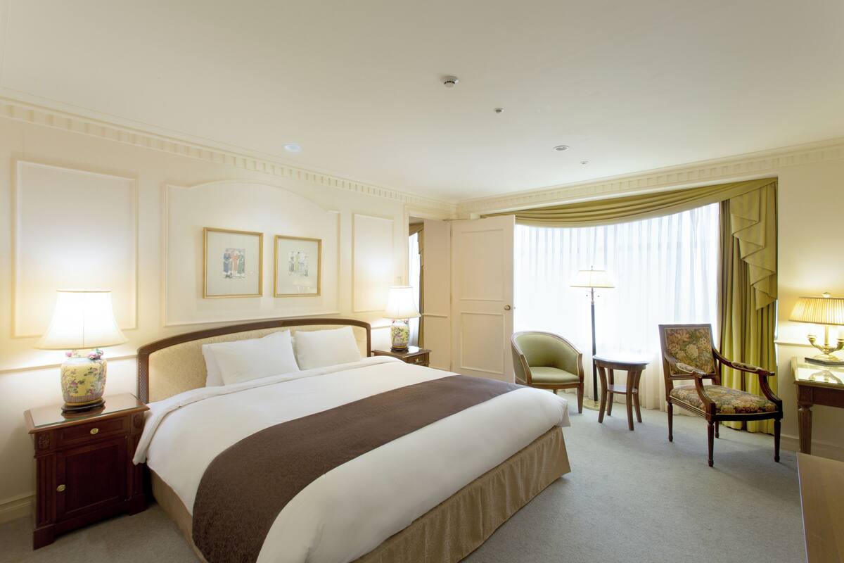 Quarto de hotel com uma cama grande, duas mesas de cabeceira com luminárias, uma cadeira, uma mesinha e uma janela com cortinas.