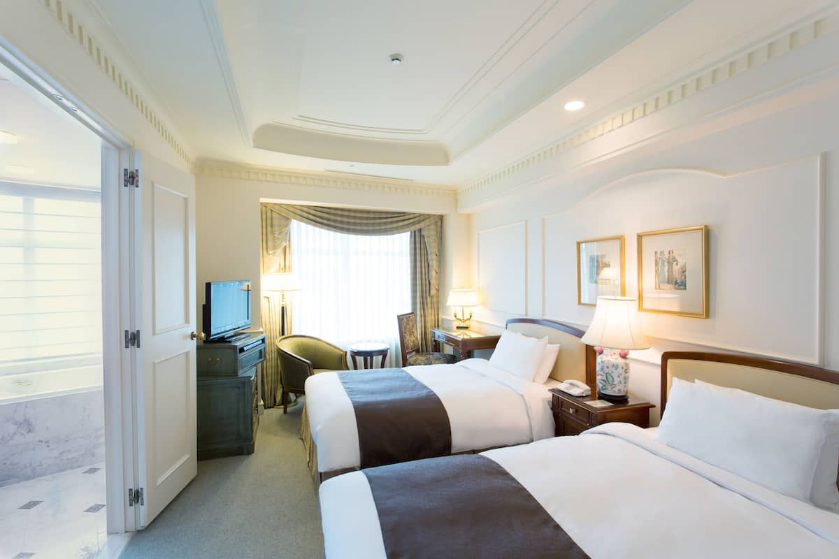 Quarto de hotel com duas camas, uma escrivaninha, uma cadeira, uma televisão e uma janela com cortinas.