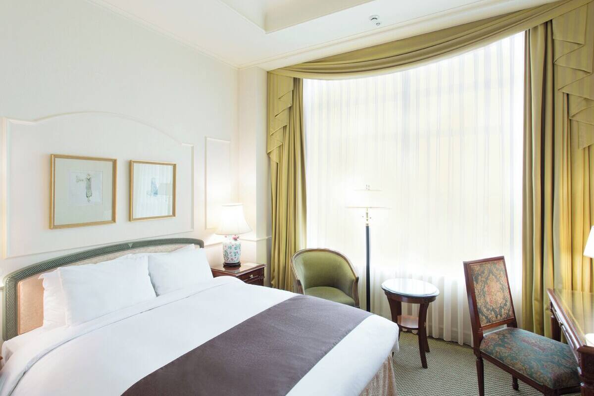 Quarto de hotel com uma cama grande, duas cadeiras, uma mesa pequena e uma janela com cortinas.