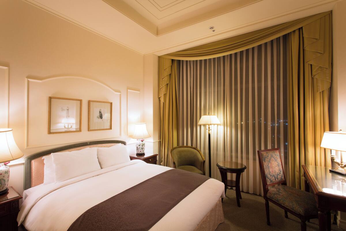 Quarto de hotel com uma cama grande, duas luminárias de cabeceira, uma cadeira, uma mesa pequena e uma janela com cortinas.