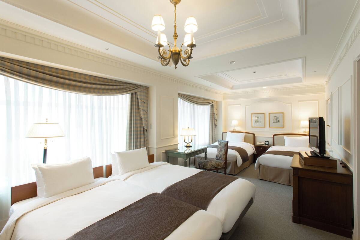 Quarto de hotel com duas camas grandes, uma escrivaninha, uma cadeira, uma televisão e um candelabro.