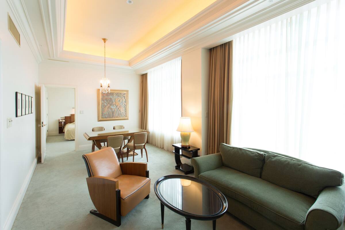 Quarto de hotel com um sofá verde, uma poltrona de couro, um espaço de refeições com mesa e cadeiras, e um candelabro.