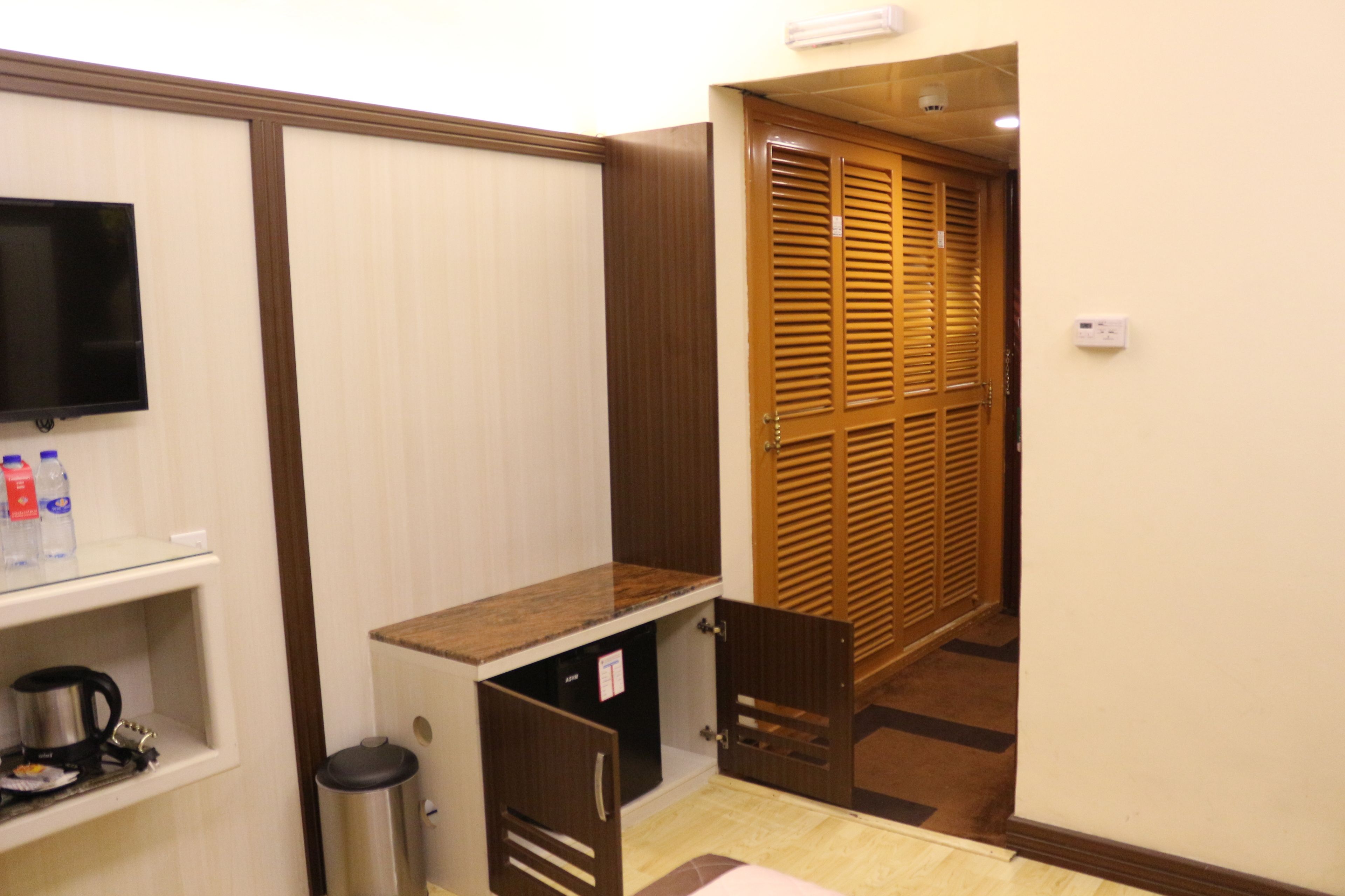 Quarto de hotel com televisão de tela plana, prateleira pequena com água engarrafada, cafeteira e guarda-roupa de madeira com portas venezianas.