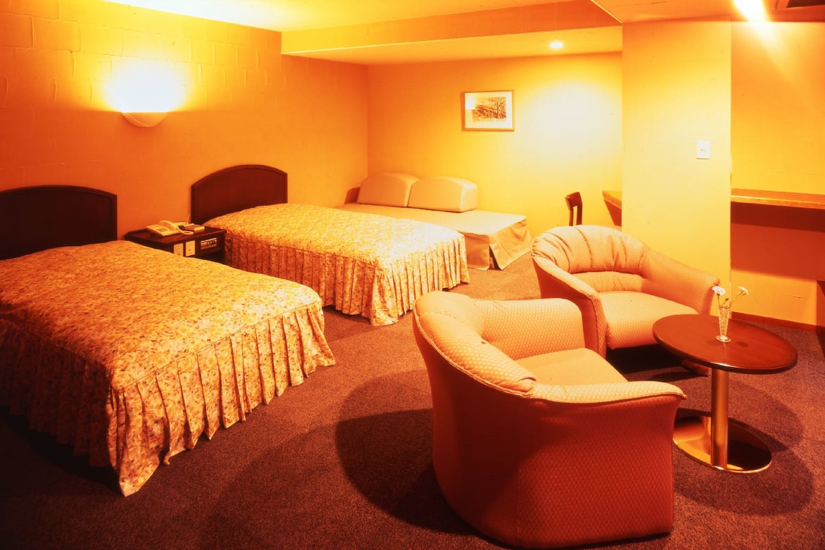 Quarto de hotel com duas camas, um sofá, uma mesa pequena e uma luminária.