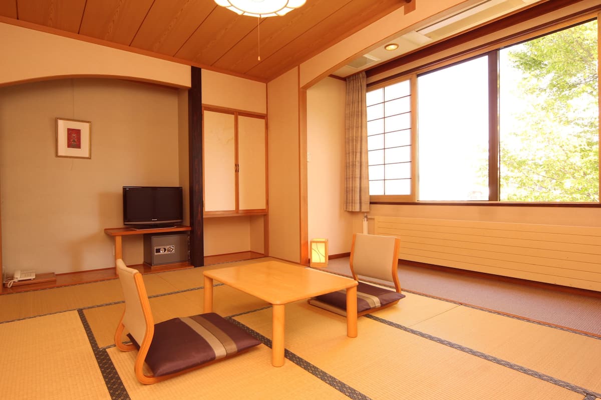 Quarto japonês tradicional com tatames, uma mesa baixa, duas cadeiras, uma televisão e uma porta deslizante.