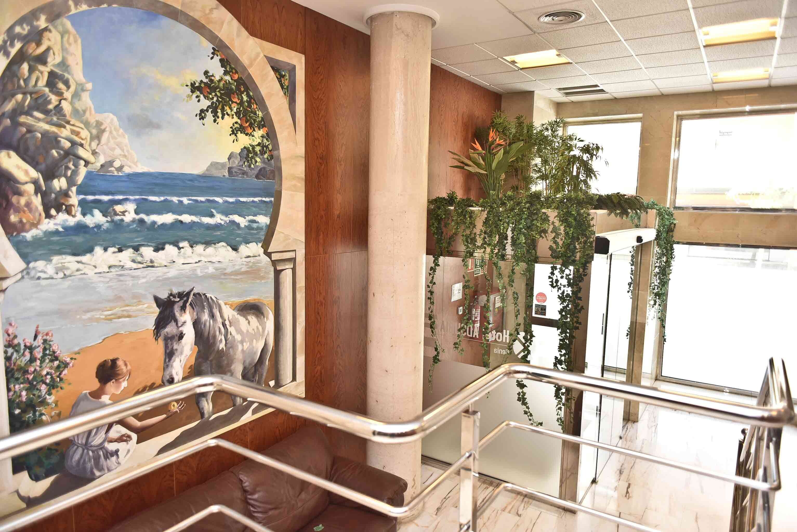Um saguão de hotel com um mural de uma cena de praia, uma escada e plantas de interior.