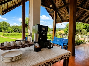 Uma área de estar coberta ao ar livre, com uma máquina de café, mesa com pratos e xícaras, e vista para um jardim exuberante.