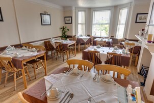 Uma sala de jantar com várias mesas preparadas para o serviço, cadeiras de madeira e uma grande janela que permite a entrada de luz natural.