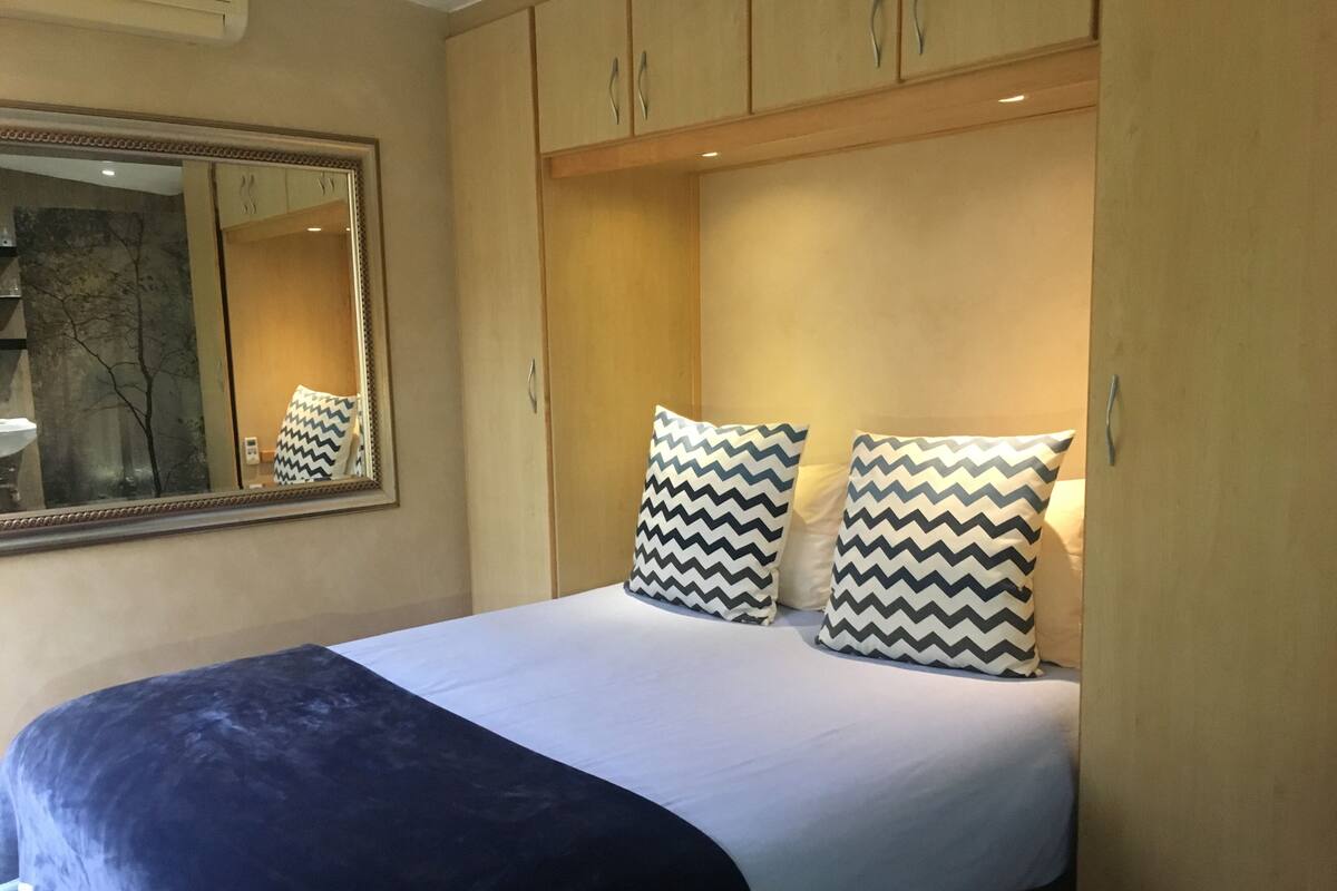 Uma cama bem arrumada com um edredom azul e duas almofadas estampadas, um espelho na parede e armários de madeira sobre a cama.