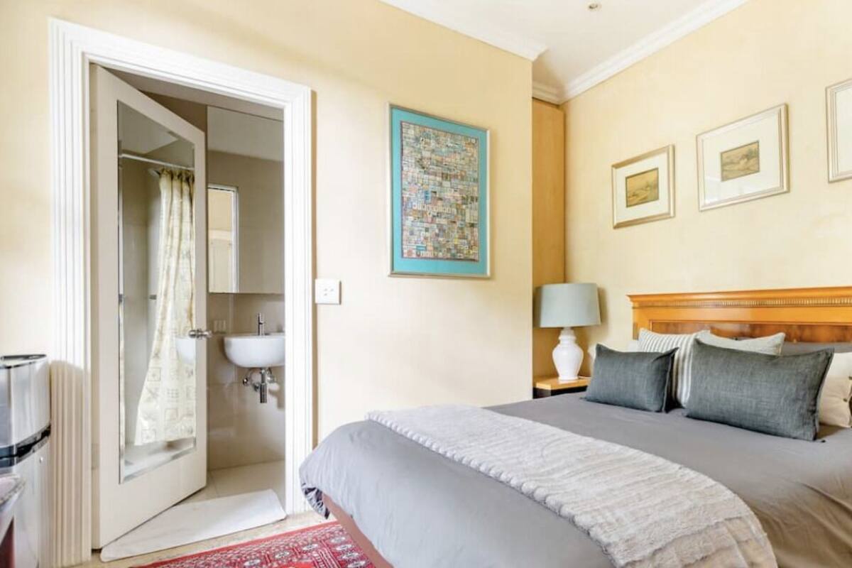 Quarto com cama, mesa de cabeceira com abajur, banheiro com cortina de chuveiro e parede com quadros.