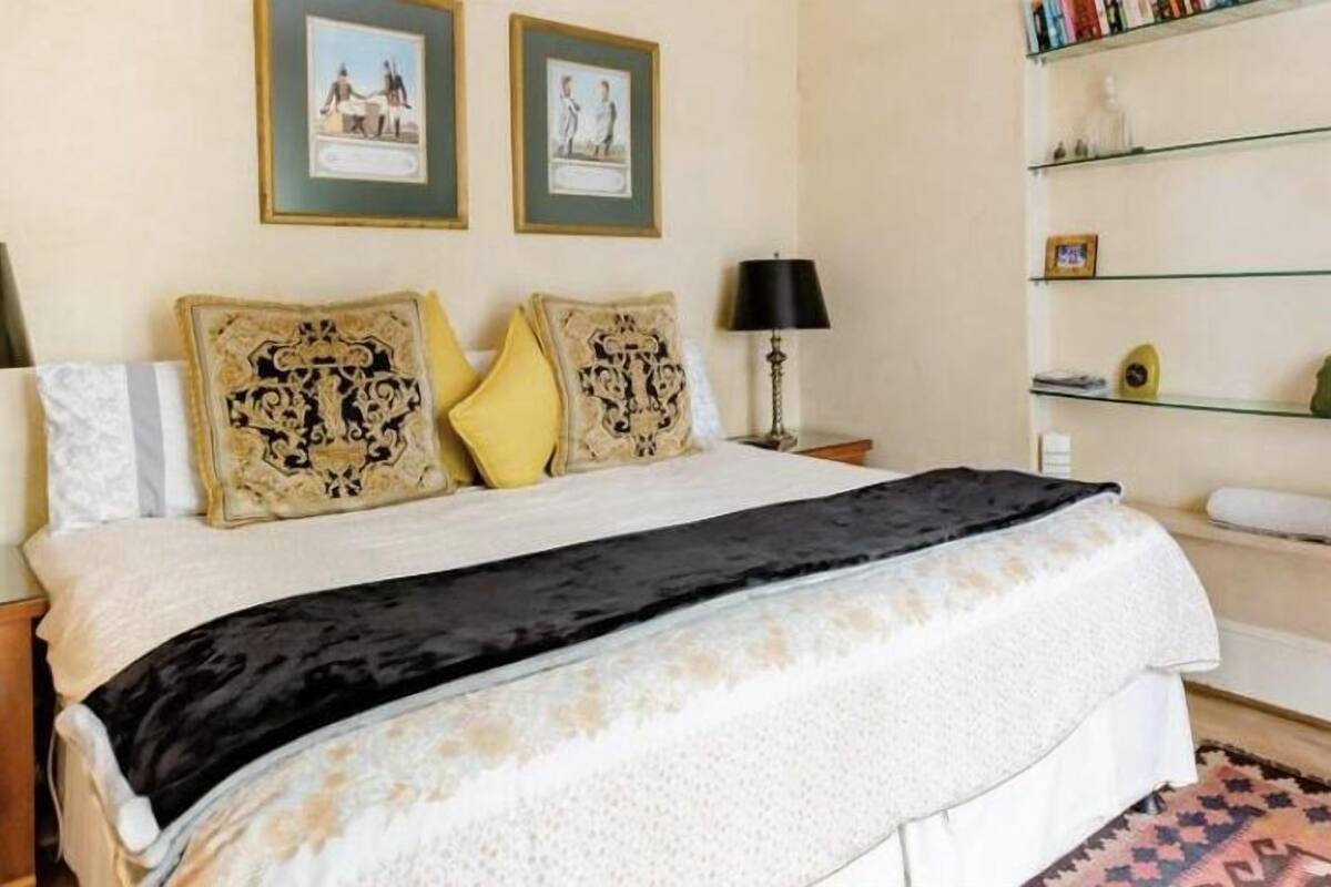 Quarto bem arrumado com cama, mesas de cabeceira, uma cadeira, estante com livros e quadros nas paredes.
