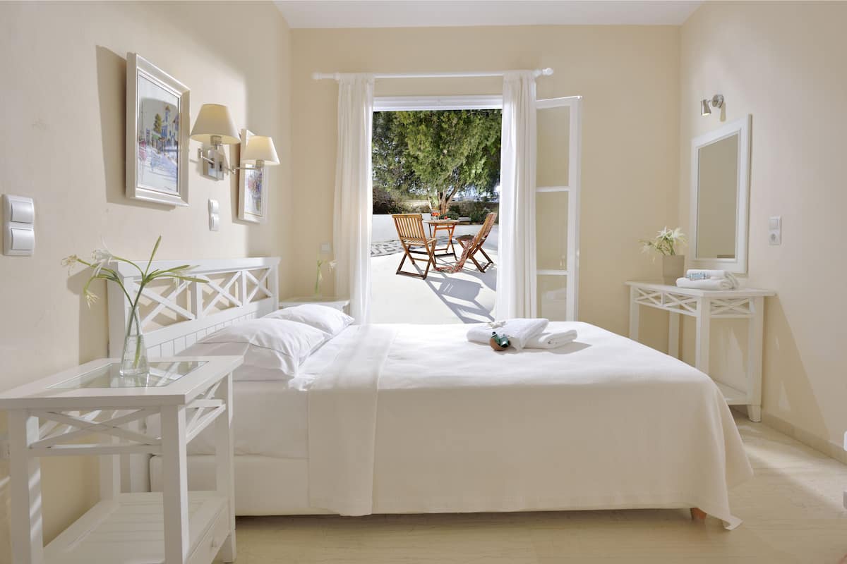 Um quarto com uma cama, mesa de cabeceira, cadeira, pequena mesa e uma janela com cortinas.