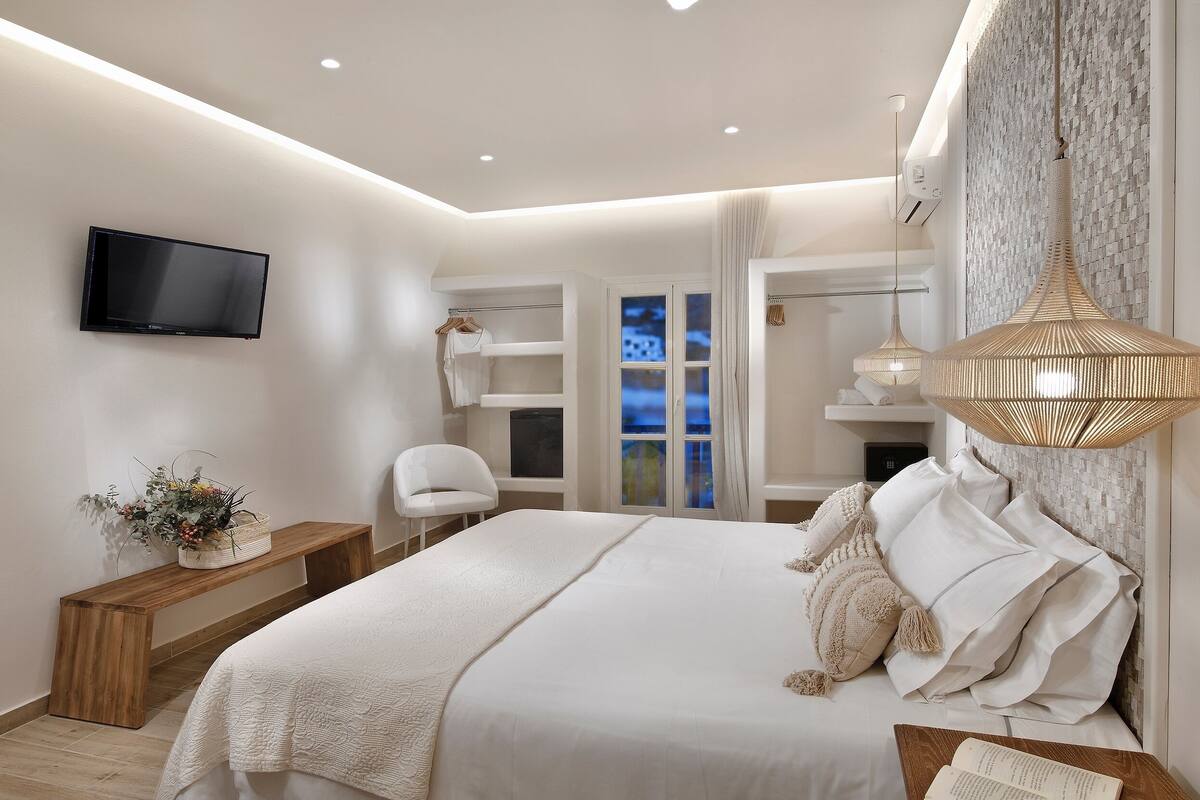 Quarto moderno com cama grande, TV fixada na parede, um banco de madeira e luminária pendente.