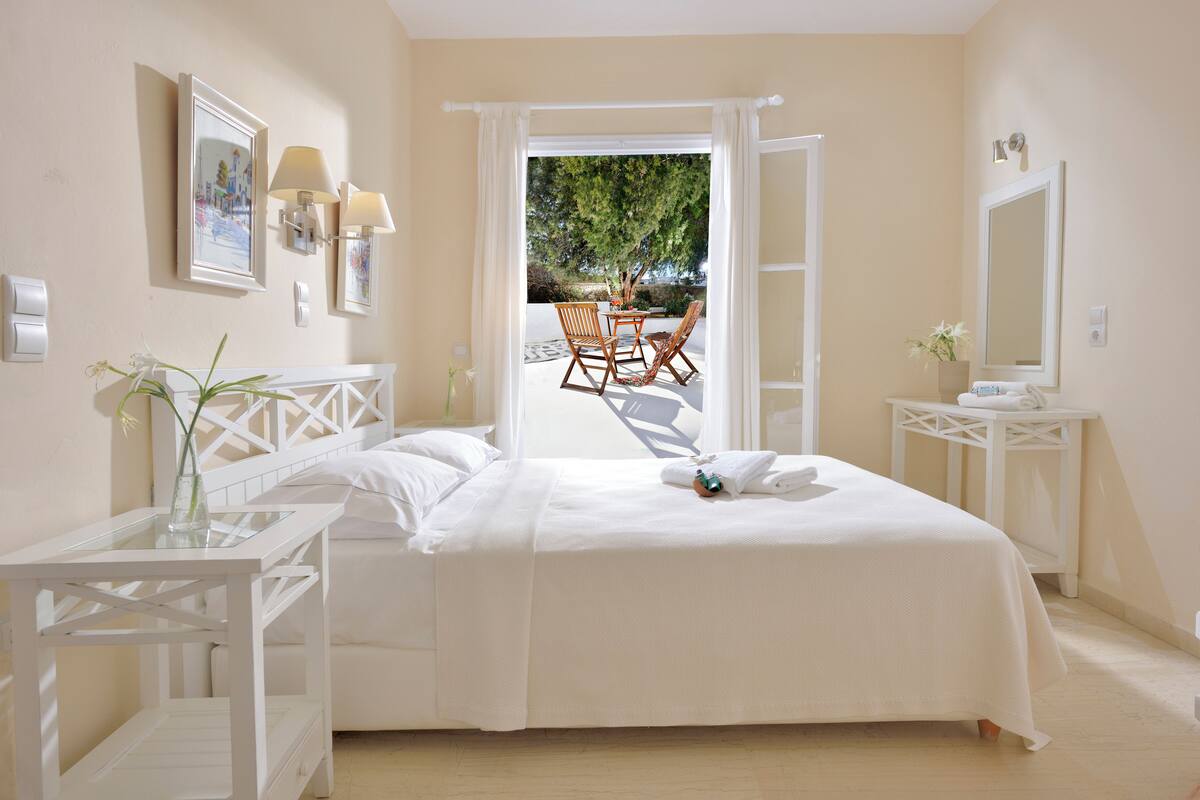 Um quarto com uma cama, mesa de cabeceira, cadeira, pequena mesa e uma janela com cortinas.
