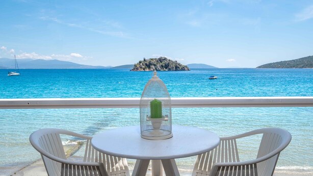 Uma varanda com mesa e cadeiras com vista para um mar azul límpido, uma pequena ilha ao longe e um guarda-corpo branco.