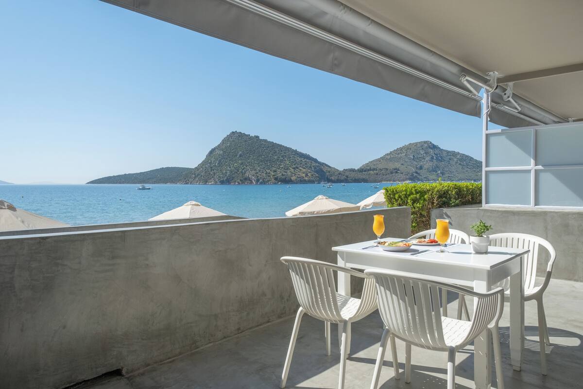Varanda com cadeiras e mesa brancas, com vista para um corpo de água com barcos e colinas ao longe.