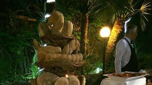 Uma mesa de buffet com diversos alimentos, incluindo uma cesta de pão em vários níveis, saladas e carnes grelhadas.