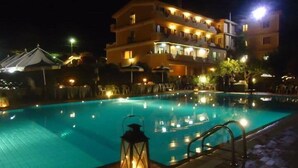 Vista noturna de um hotel com piscina, área de estar externa e fachada bem iluminada.