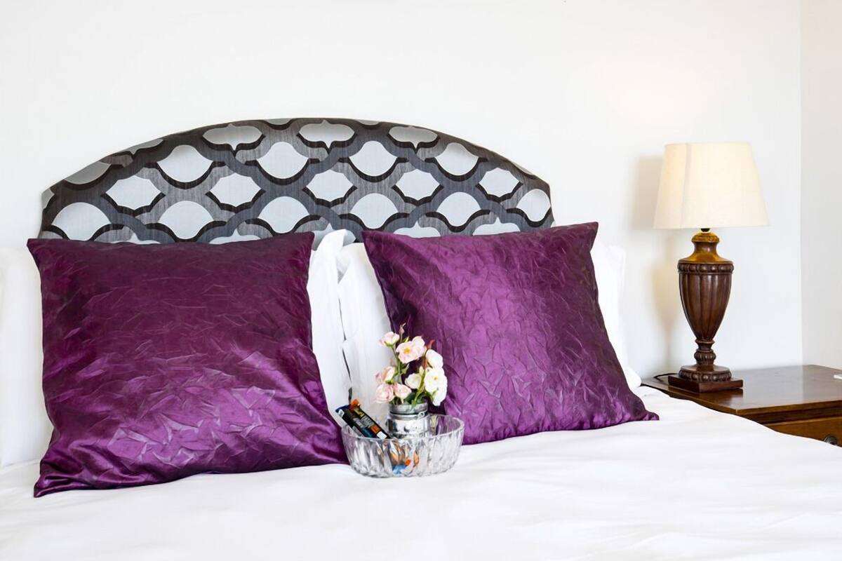Uma cama com dois travesseiros roxos, um travesseiro branco e um vaso com flores.
