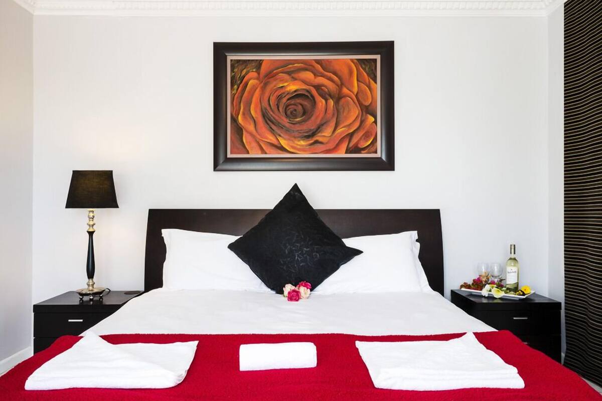 Quarto de hotel com cama grande, roupa de cama branca, cabeceira preta, pintura emoldurada de uma rosa, mesa de cabeceira com abajur e bandeja com garrafa e flores.