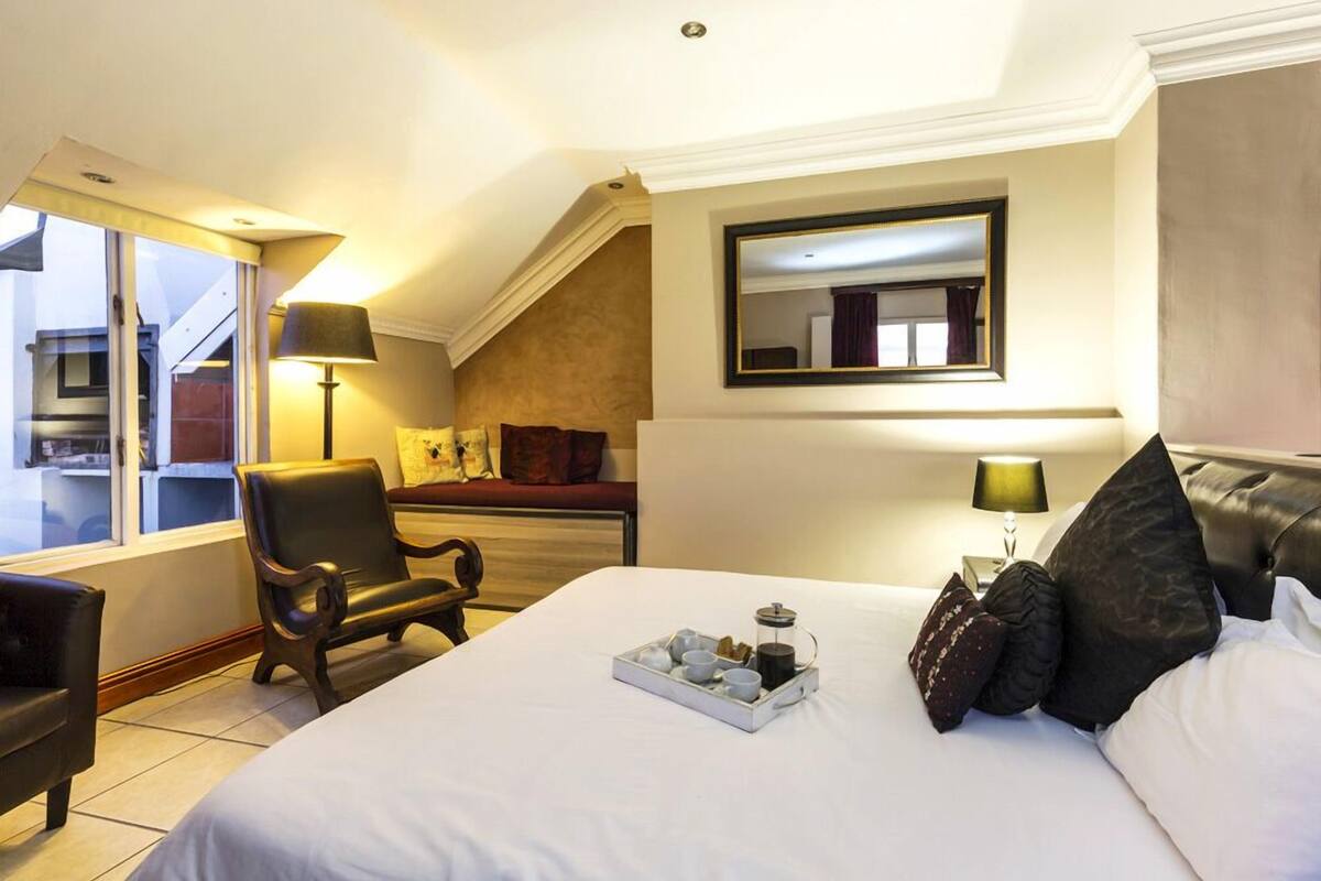 Quarto de hotel com uma cama grande, um espaço de estar com uma cadeira e uma mesa, uma janela com vista e um espelho na parede.
