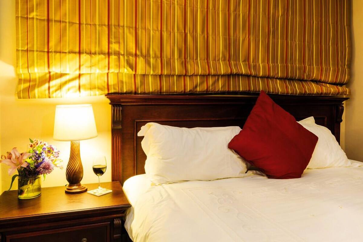 Uma cama bem arrumada com lençóis brancos e um travesseiro vermelho, uma mesinha de cabeceira com um abajur e um vaso de flores, e uma cabeceira de madeira.