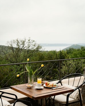 Uma mesa de madeira com um copo de suco de laranja, uma tigela de frutas e um prato de comida numa varanda com vista para árvores e um corpo de água.