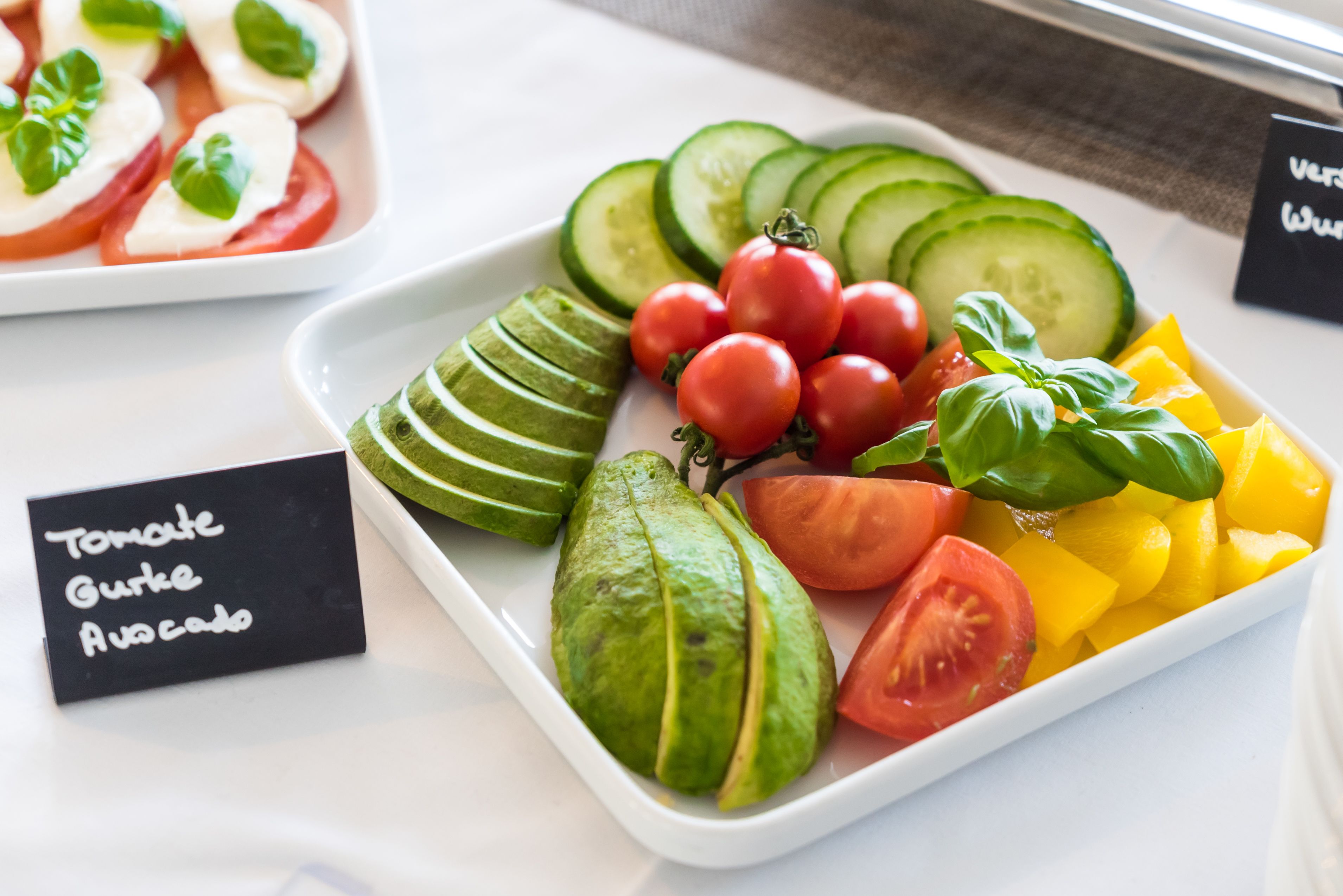 Um prato branco com fatias de abacate, tomates cereja, fatias de pepino e pedaços de manga amarela, rotulado como 'Tomate Gurke Avocado'.