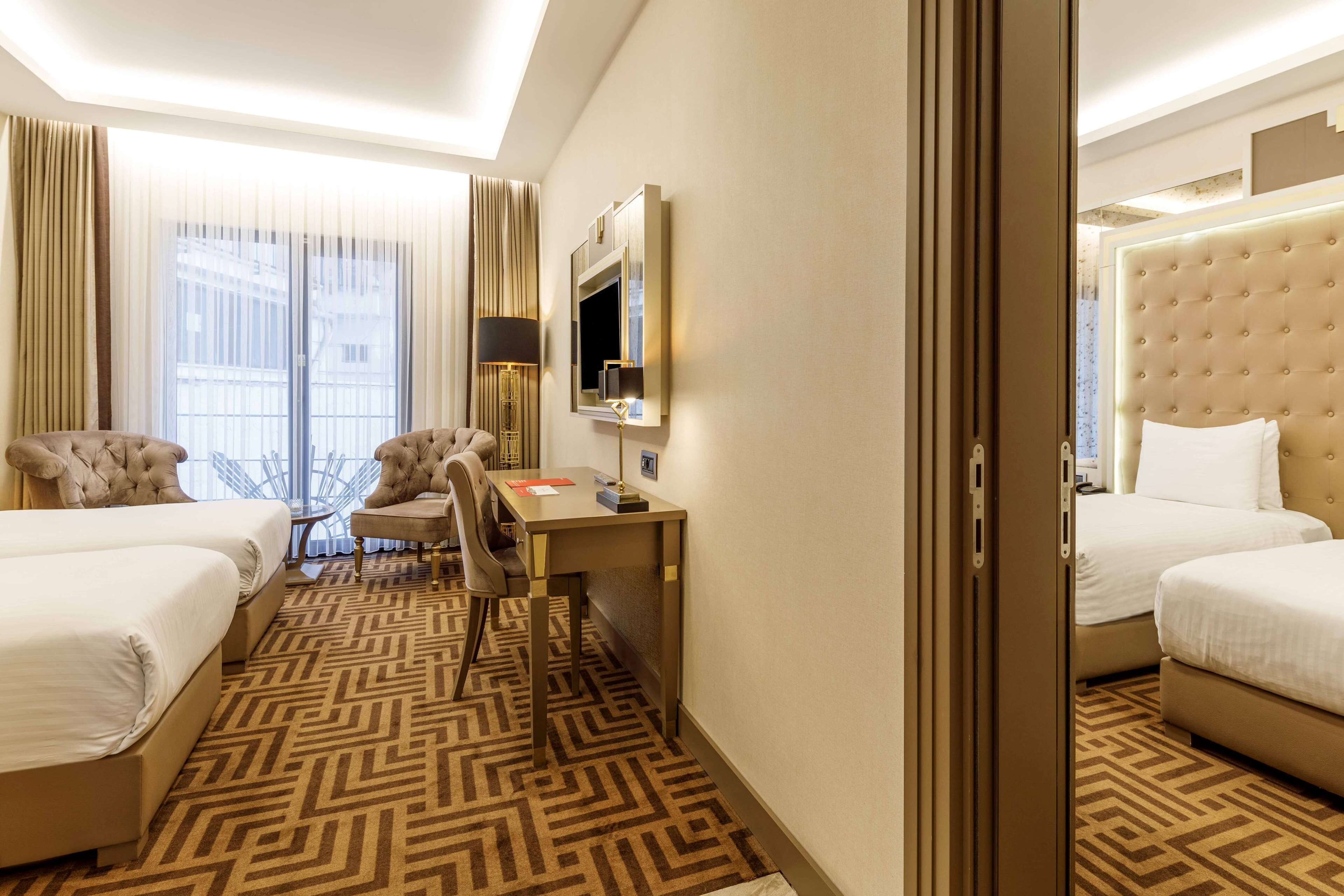 Quarto de hotel com duas camas, uma escrivaninha, uma cadeira e uma janela grande com cortinas.