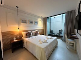 Quarto de hotel com uma cama grande, mesa de cabeceira, escrivaninha com cadeira, TV na parede e porta de vidro corrediça para uma varanda.