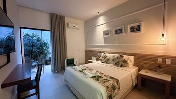 Quarto de hotel moderno com cama, escrivaninha, cadeira e vista para vegetação através de uma porta deslizante.