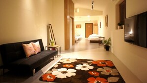 Uma sala de estar moderna com um sofá, uma mesa de centro e um tapete floral. Há um corredor que leva a um quarto com uma cama e uma TV fixada na parede.