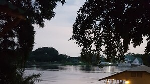 Um banco de madeira e uma mesa perto de um lago calmo ao entardecer, com uma casa e um cais ao fundo.