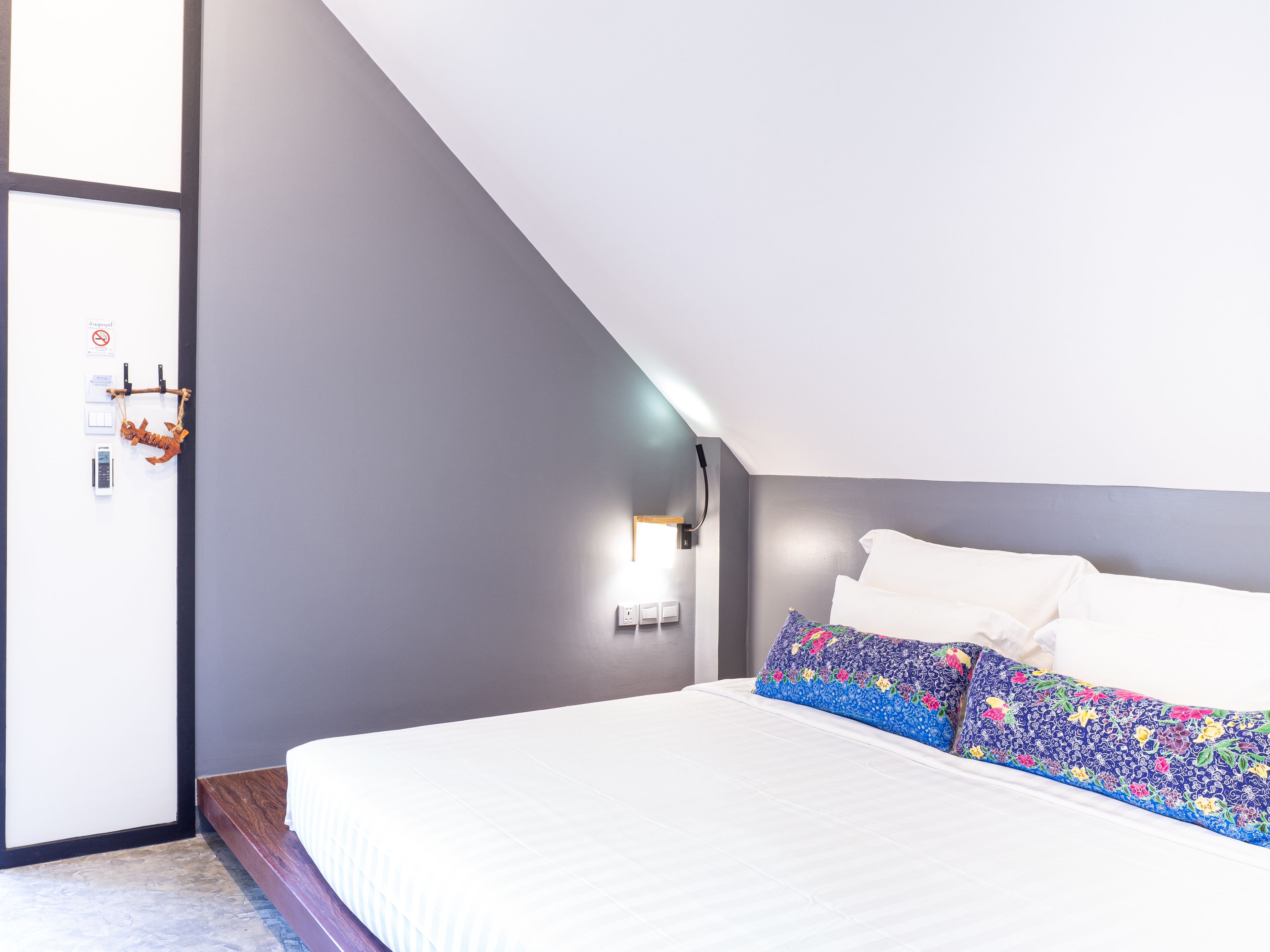 Quarto moderno com teto inclinado, cama com roupa de cama branca, almofadas florais coloridas e luminária fixada na parede.