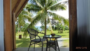 Um deck de madeira com duas cadeiras de vime e uma mesa pequena, com vista para um jardim tropical com palmeiras e o oceano.