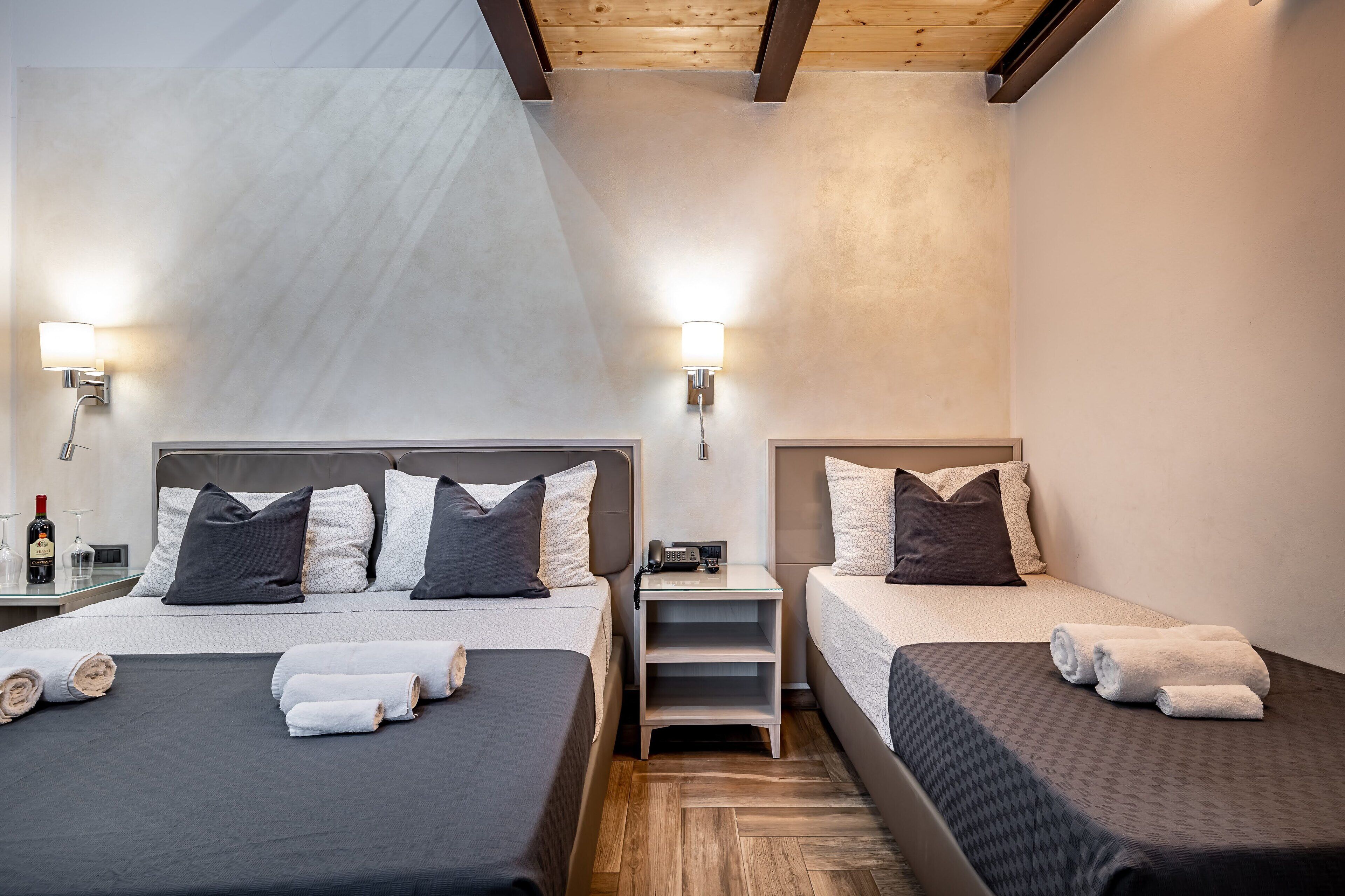 Quarto de hotel com duas camas, cada uma com um travesseiro, uma mesa de cabeceira com luminária e uma pequena prateleira.