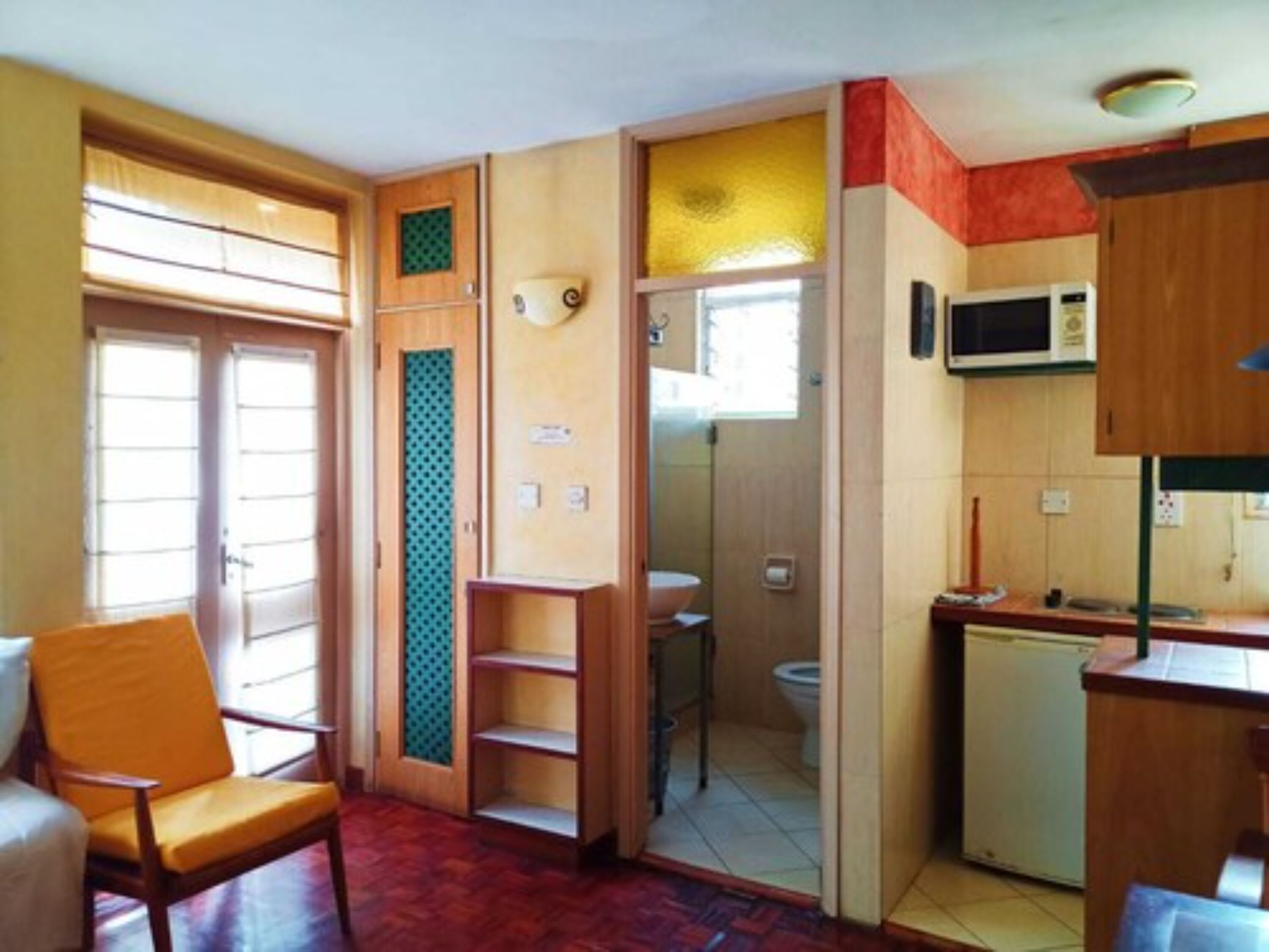Uma cozinha compacta com micro-ondas, um pequeno frigorífico e uma pia. Há uma cadeira e uma estante no quarto.