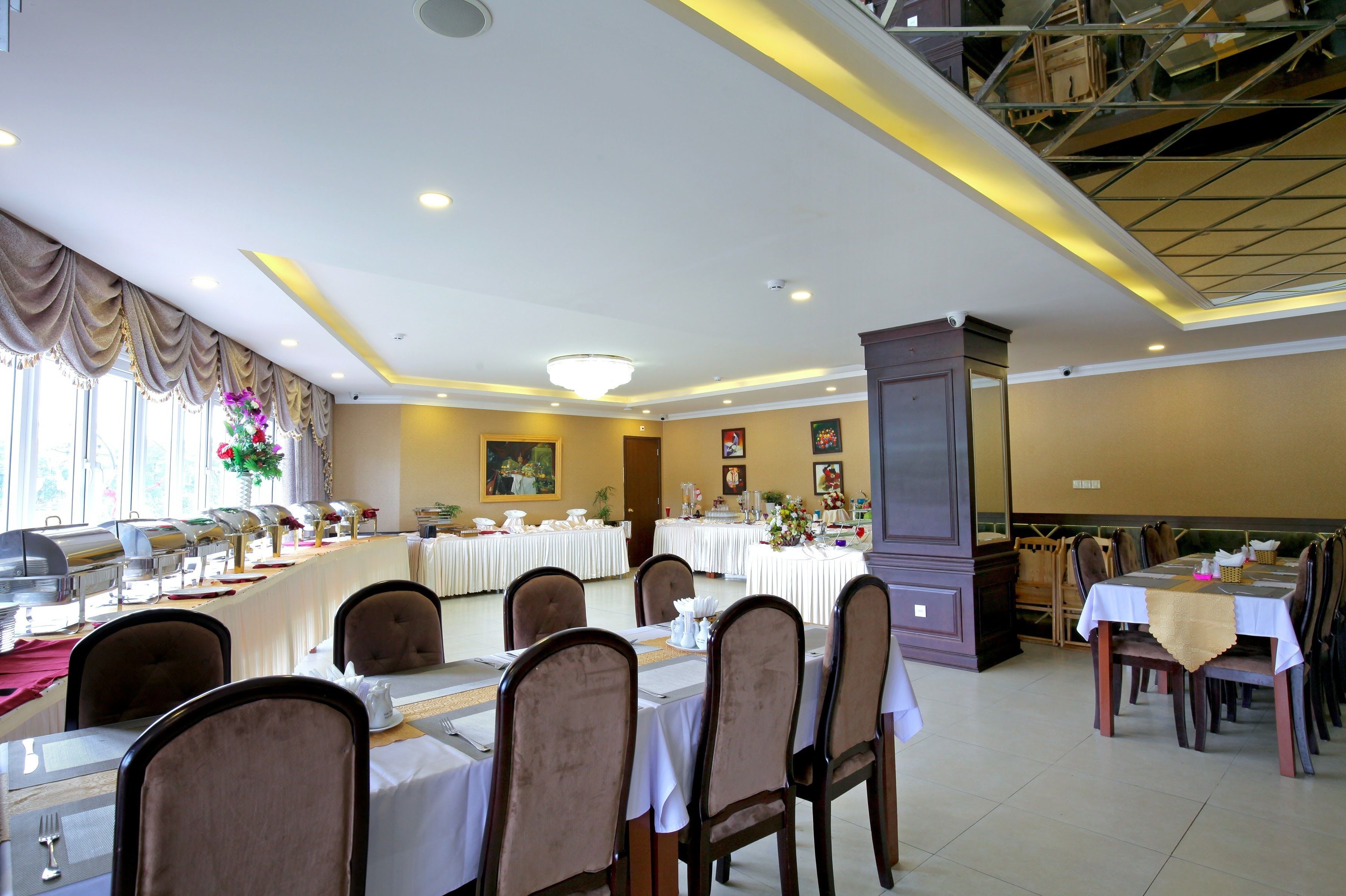 Uma sala de jantar com várias mesas e cadeiras, um serviço de buffet e uma grande janela com cortinas.