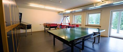 Quarto com mesa de pingue-pongue, mesa de futebol de mesa e mesa de tênis. Janelas oferecem vista para árvores lá fora.