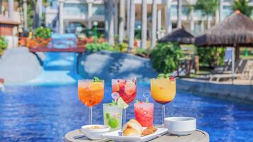 Mesa à beira da piscina com bebidas e petiscos, um toboágua e um guarda-sol de palha em um ambiente de resort.