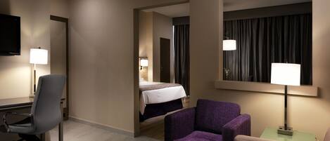 Quarto de hotel com escrivaninha, duas cadeiras, uma cama, televisão e janela com cortinas.