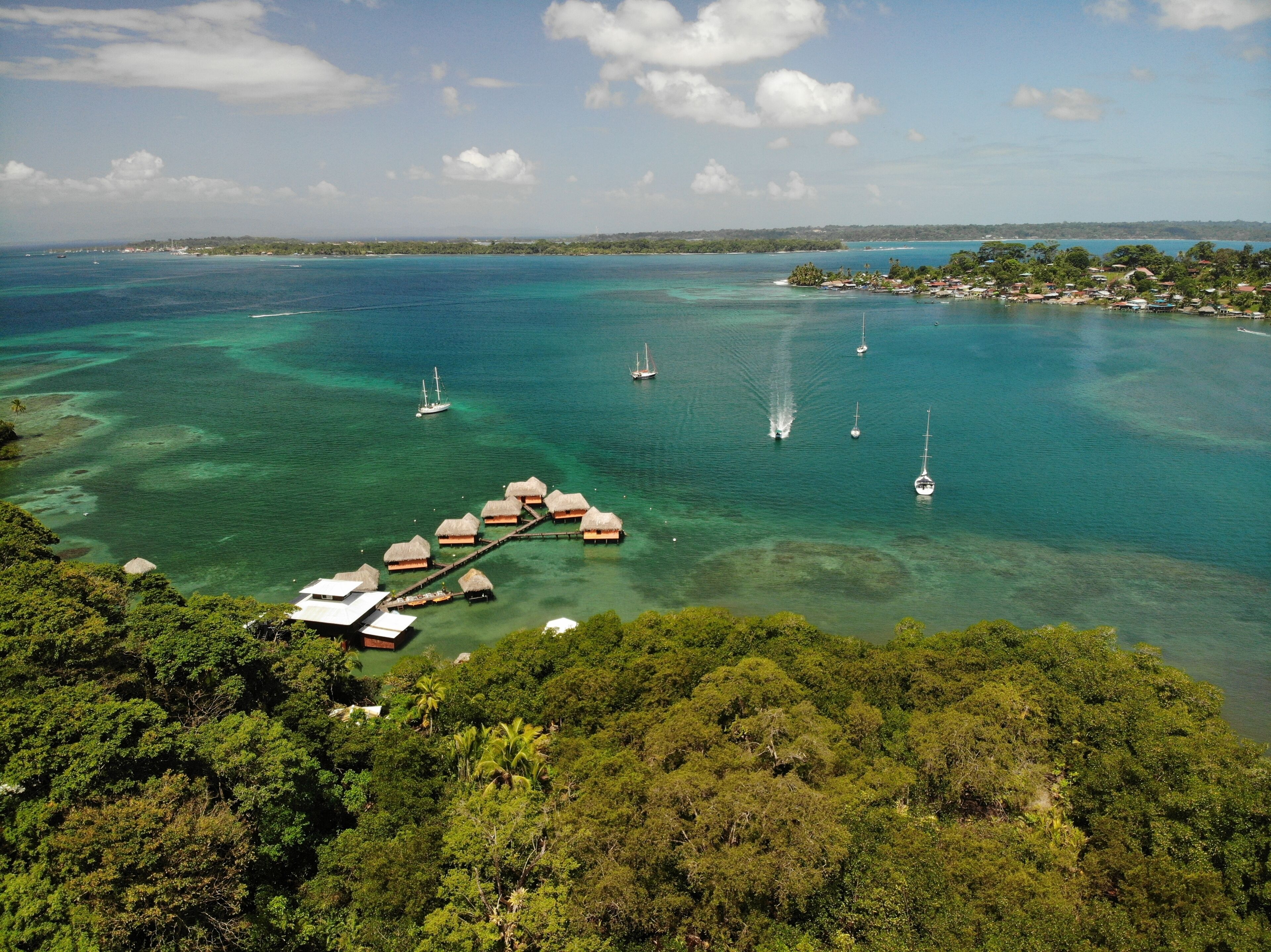 Vista aérea de uma ilha tropical com águas turquesa cristalinas, bangalôs sobre a água e um pequeno povoado com casas e barcos.